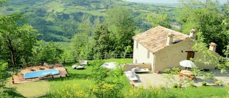 Uma casa de pedra com telhado de telhas, rodeada por uma paisagem verde exuberante e área de piscina com espreguiçadeiras.