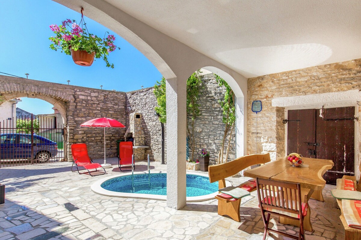 Uma área à beira da piscina com piso de pedra, uma mesa de madeira, cadeiras vermelhas e um vaso de flores pendurado.