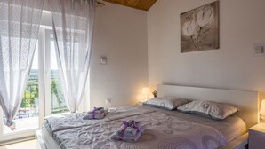 Quarto com uma cama grande, duas mesas de cabeceira, uma janela com cortinas leves e um quadro na parede.