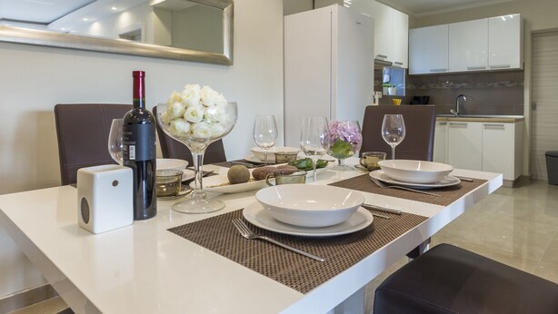 Uma sala de jantar moderna com uma mesa branca posta para dois, incluindo uma garrafa de vinho, copos e um arranjo floral branco.