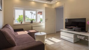 Uma sala de estar moderna com um sofá marrom, uma TV sobre um móvel branco, uma janela com plantas e uma porta.