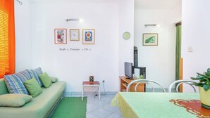 Sala de estar com sofá, mesinha, televisão e tapete verde. Quadros emoldurados na parede e uma frase motivacional.