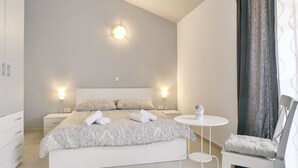 Um quarto moderno com uma cama grande, mesas de cabeceira, uma pequena mesa redonda, uma cadeira e uma janela com cortinas.