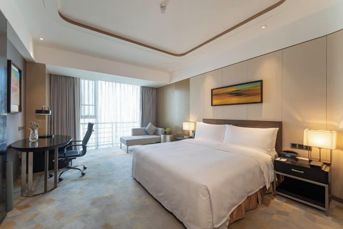 Quarto de hotel com uma cama grande, escrivaninha, cadeira, mesa de cabeceira e abajur.