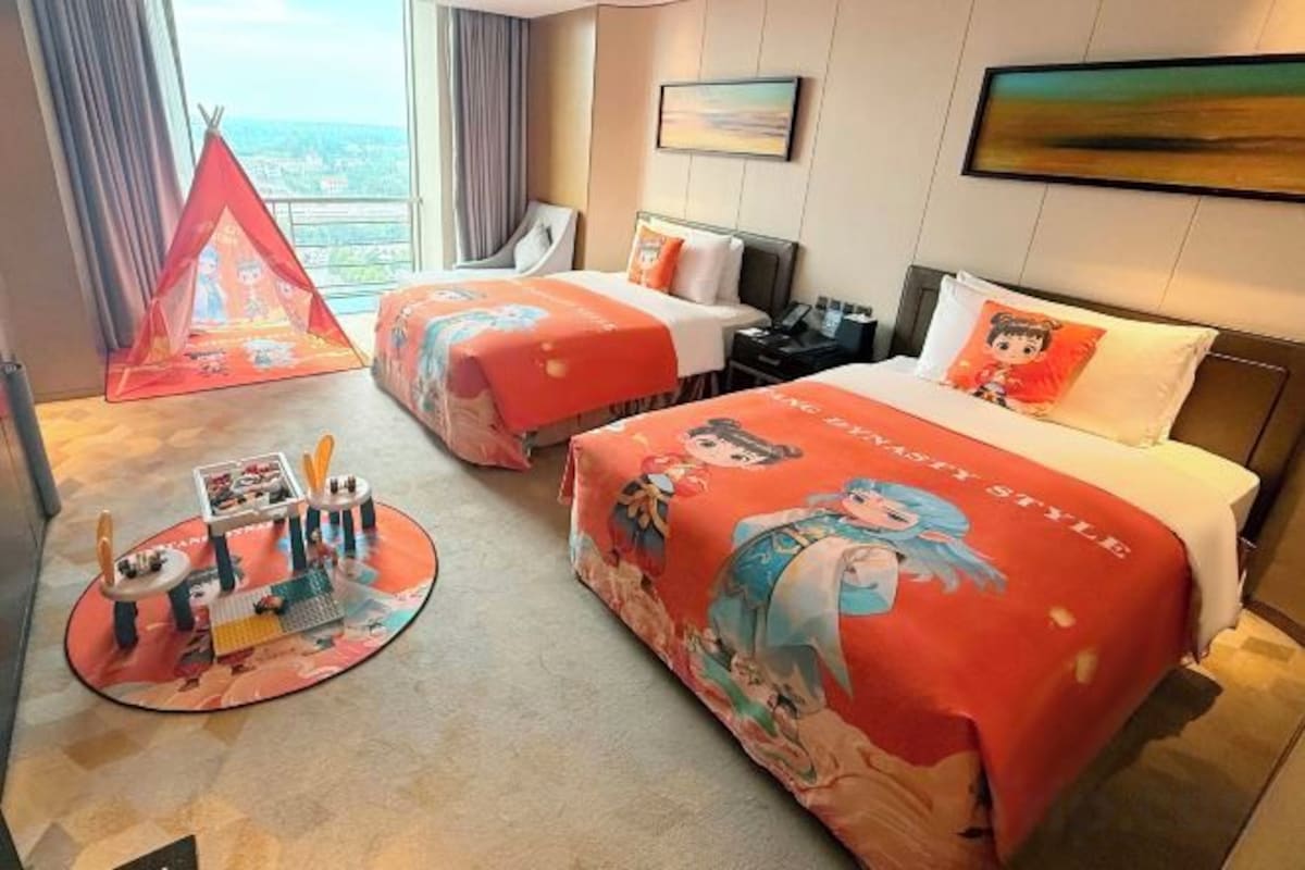 Quarto de hotel com duas camas, colcha de cama com tema vermelho e branco, uma pequena mesa com bebida e petiscos, uma cadeira e uma janela com vista.