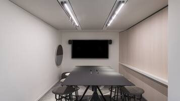 Uma sala de conferências com uma mesa grande, quatro cadeiras, uma tela de projetor e uma TV fixada na parede.
