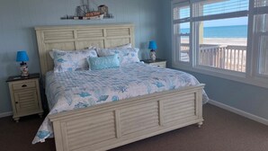 Quarto com cama, duas mesas de cabeceira, ventilador de teto, janela com vista para a praia e um modelo decorativo de navio na parede.