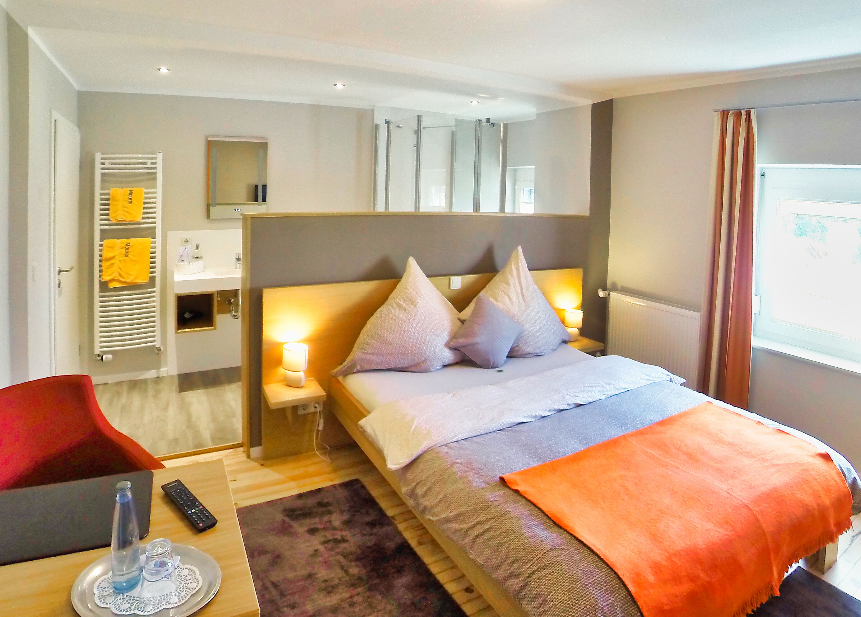 Quarto de hotel com cama, mesas de cabeceira, uma cadeira, escrivaninha, janela e banheiro com porta-toalhas.