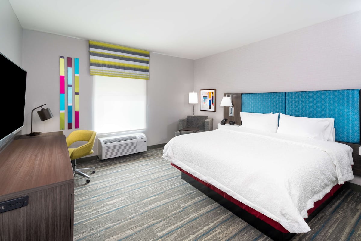 Quarto de hotel com cama grande, escrivaninha, cadeira, televisão e janela com persianas.