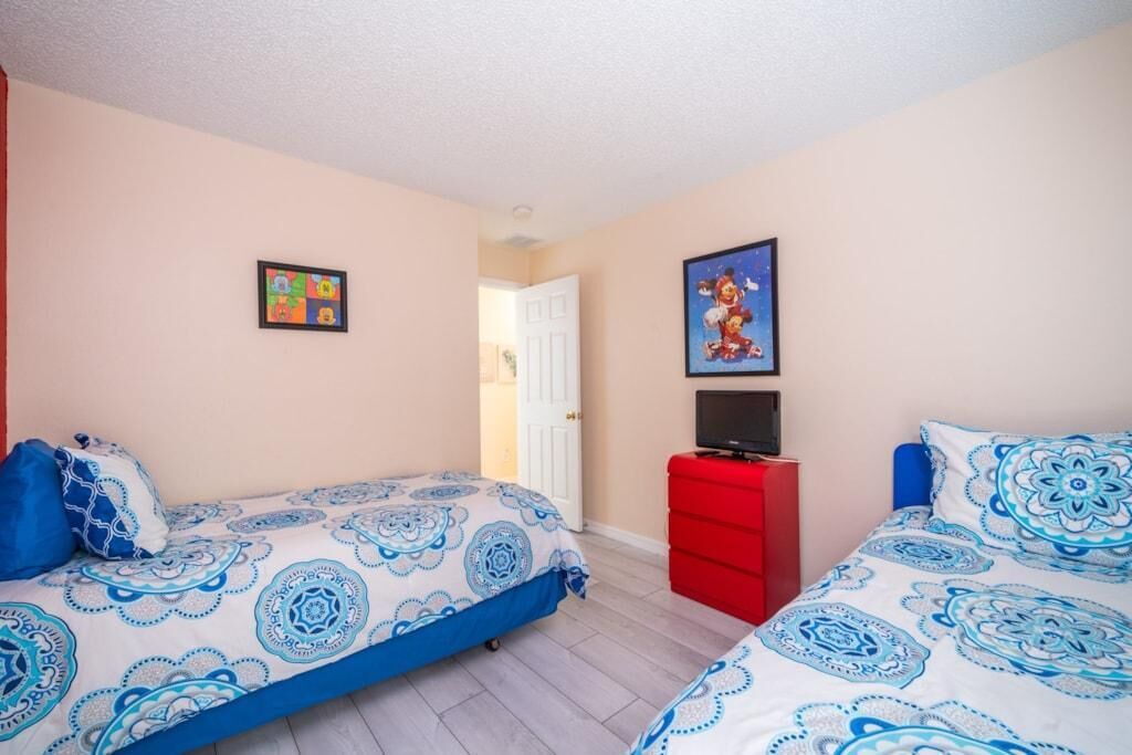 Quarto com duas camas, uma cômoda vermelha, televisão e duas obras de arte nas paredes.