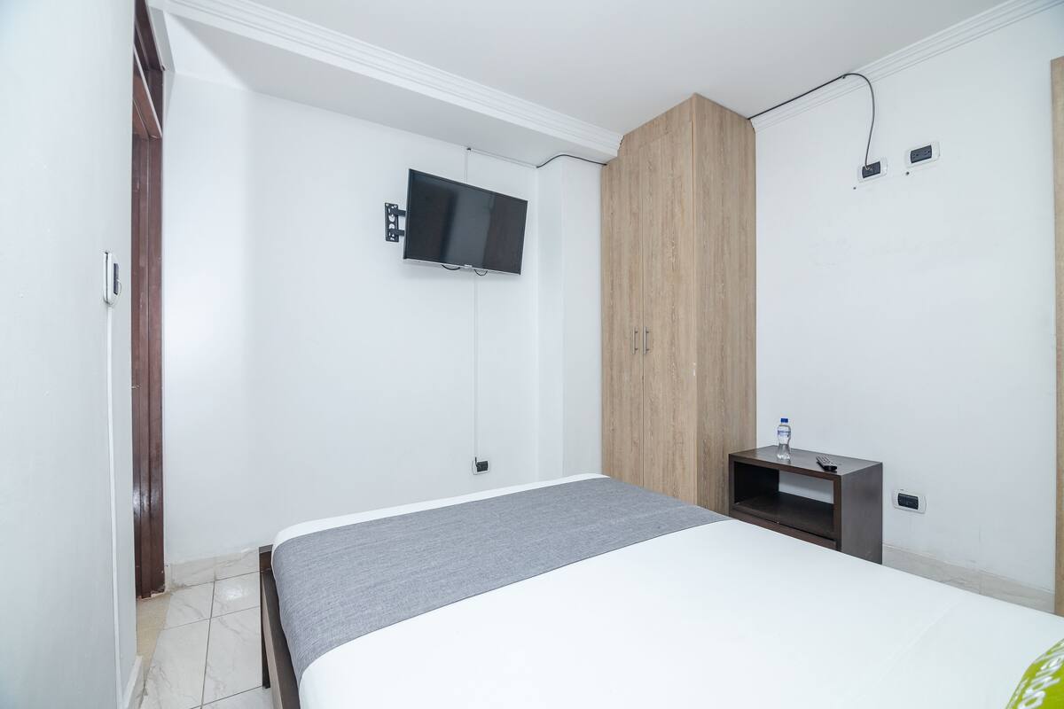 Quarto com cama, televisão presa à parede, armário de madeira e uma pequena mesa de cabeceira com uma garrafa de água.