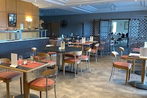 Restaurante com várias mesas e cadeiras, um balcão com cardápio e um relógio na parede.