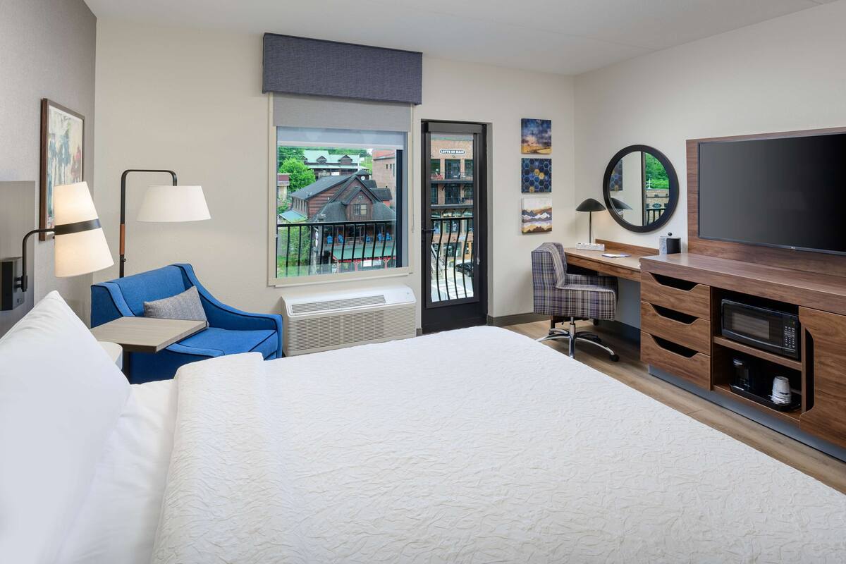 Quarto de hotel com uma cama grande, uma escrivaninha com cadeira, televisão e janela com vista para edifícios.