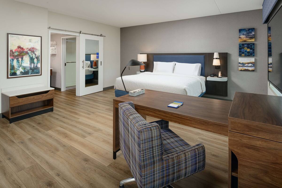 Quarto de hotel com cama, escrivaninha, cadeira, guarda-roupa e um quadro na parede.
