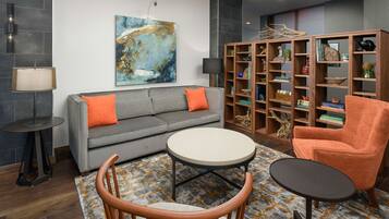 Uma sala de estar moderna com um sofá cinzento, uma poltrona laranja e mesas redondas. Uma grande pintura abstrata e uma estante de madeira com objetos decorativos também são visíveis.