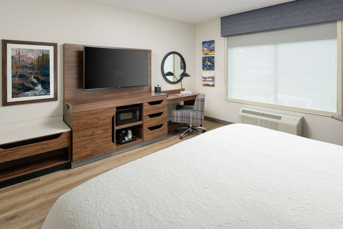Quarto de hotel moderno com uma cama grande, televisão de tela plana, móvel de entretenimento em madeira, uma escrivaninha com cadeira e obras de arte nas paredes.