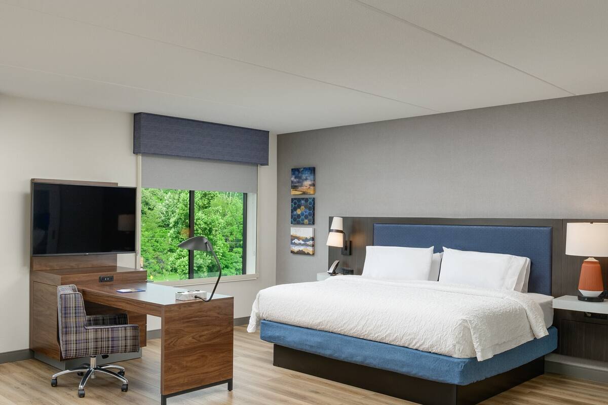 Quarto de hotel com cama, escrivaninha, cadeira, televisão e obras de arte nas paredes.