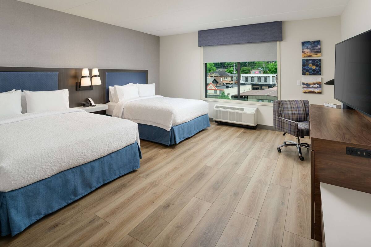 Quarto de hotel com duas camas, uma escrivaninha, uma cadeira, televisão e janela com vista para edifícios.