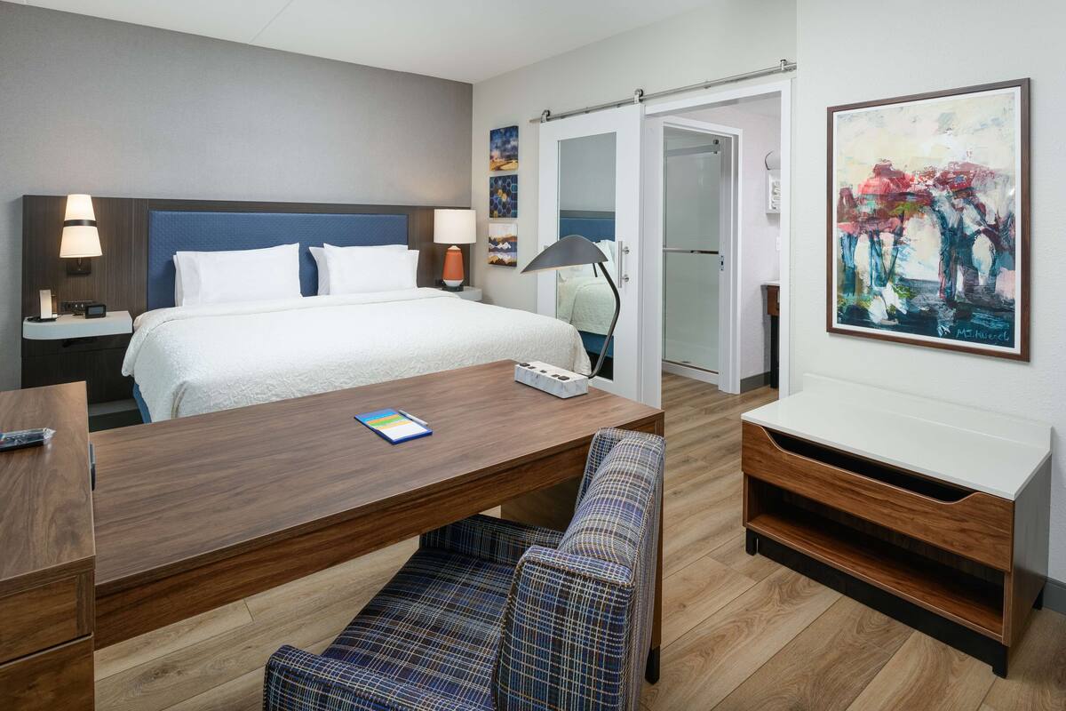 Quarto de hotel com uma cama, uma escrivaninha, uma cadeira, um abajur e um quadro na parede.