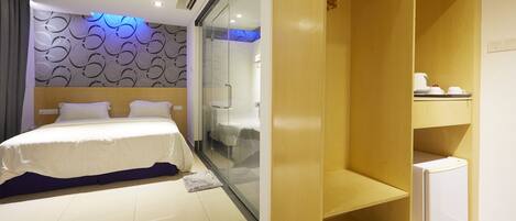Quarto de hotel moderno com cama, box de vidro, guarda-roupa de madeira e um pequeno frigorífico branco.