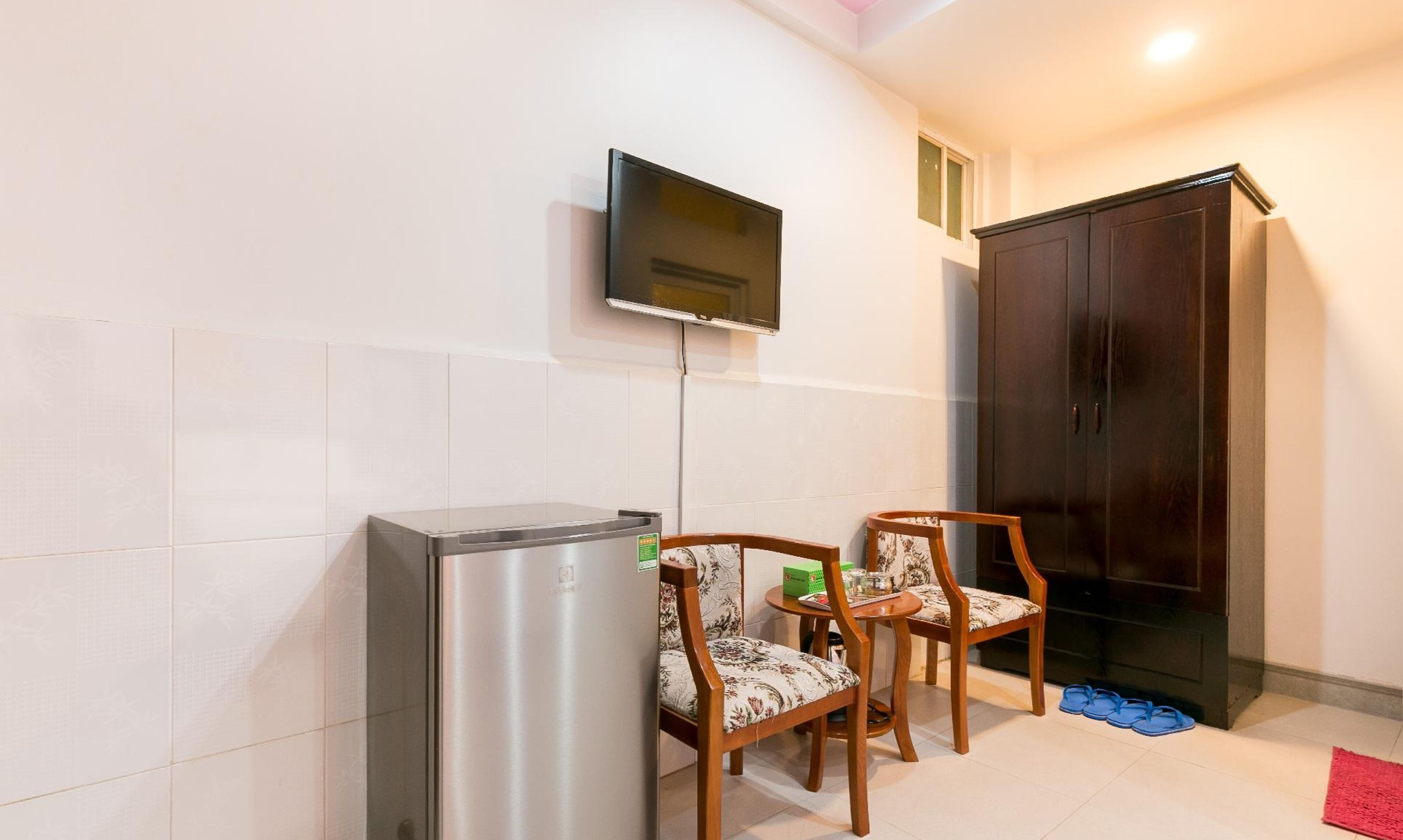 Quarto compacto com geladeira, duas cadeiras, uma mesa pequena com uma caixa, TV fixada na parede e um guarda-roupa escuro.