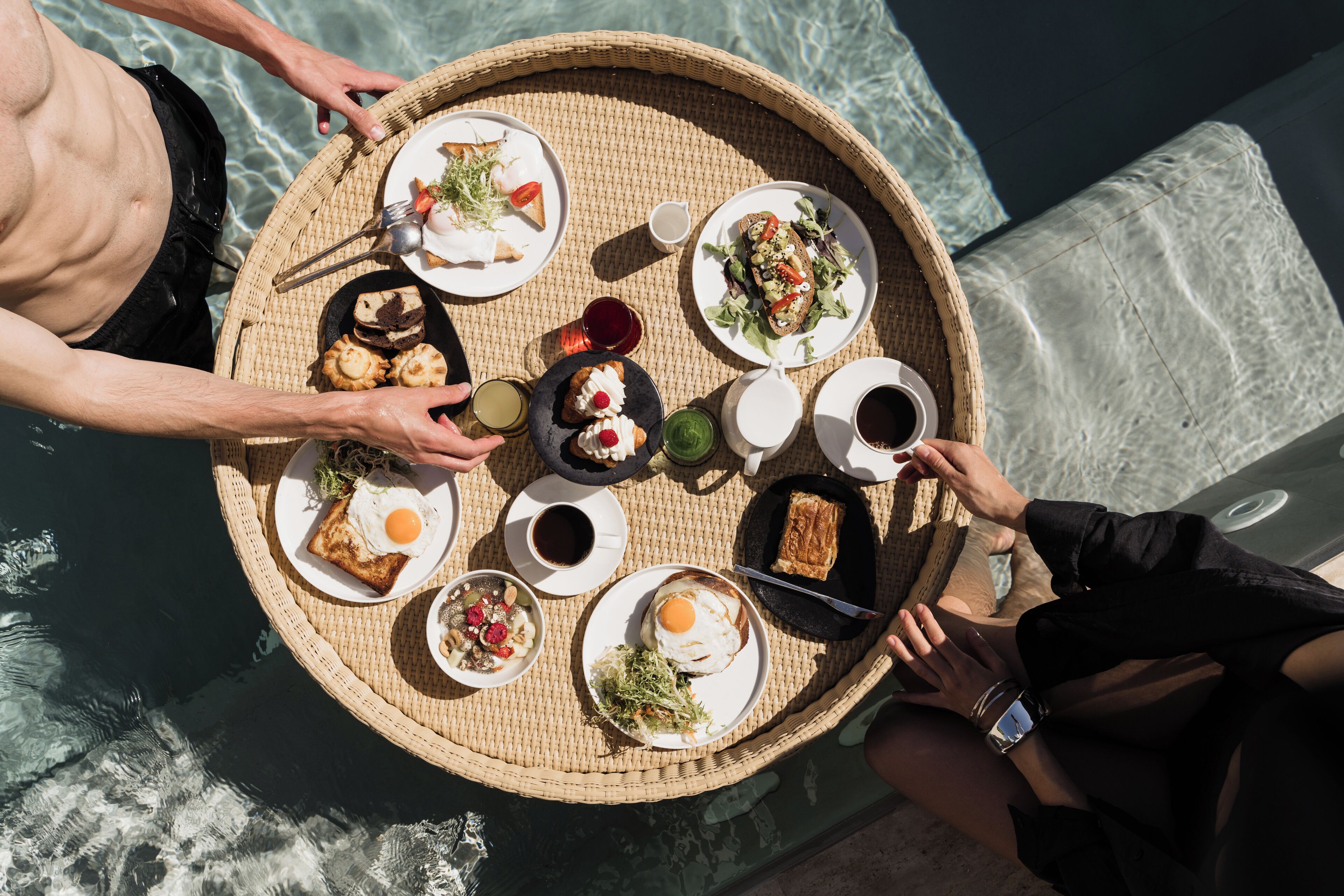 Uma bandeja de vime com diversos itens de pequeno-almoço e bebidas, colocada sobre uma mesa de vidro junto a uma piscina.