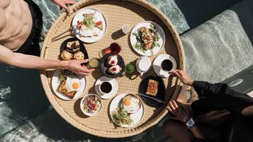 Uma bandeja de vime com diversos itens de pequeno-almoço e bebidas, colocada sobre uma mesa de vidro junto a uma piscina.