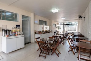 Uma sala de jantar espaçosa, com várias mesas e cadeiras, balcão branco com eletrodomésticos e grandes janelas que proporcionam luz natural.