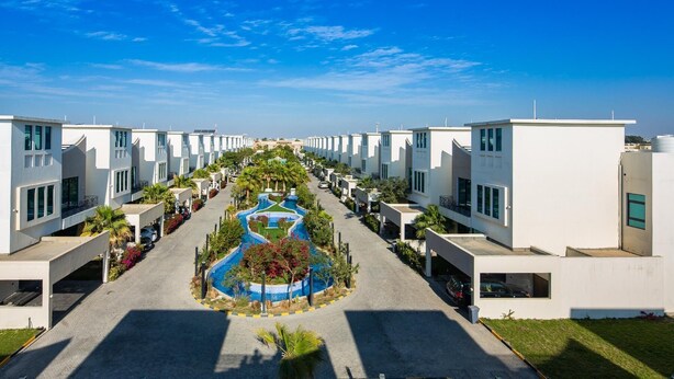 Uma área residencial moderna com edifícios brancos, um caminho central com paisagismo e um céu azul claro.