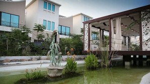 Um edifício moderno com área externa coberta, um pequeno lago com plantas aquáticas e uma escultura.