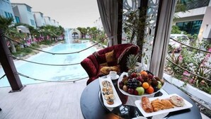 Mesa de jantar com diversos doces, frutas e café, com vista para uma piscina e área ajardinada.