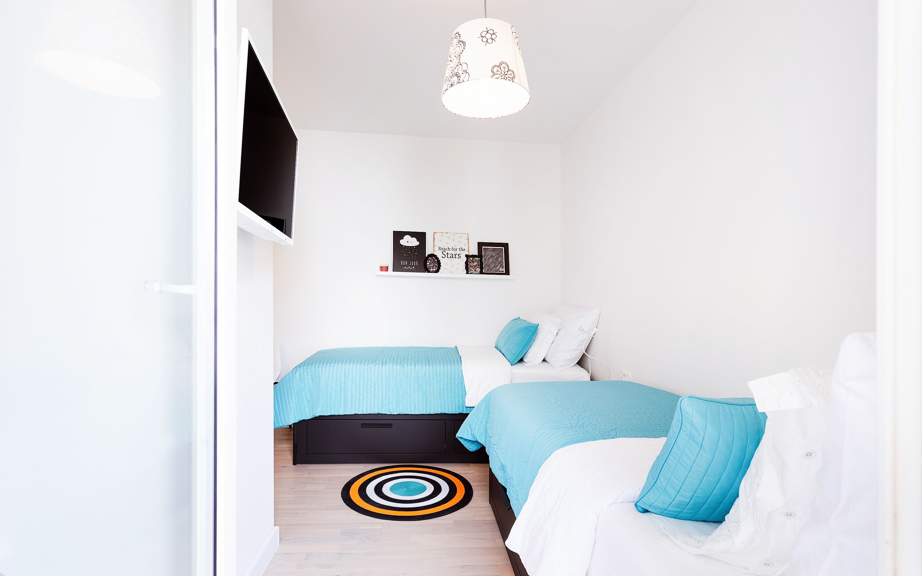 Quarto com duas camas, TV fixada na parede e luminária no teto.