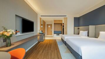 Quarto de hotel moderno com uma cama grande, televisão na parede, escrivaninha com um vaso de flores e vista para o corredor.