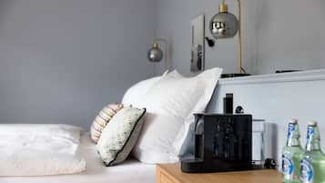 Cama bem arrumada com lençóis brancos e almofadas estampadas, mesa de cabeceira com um alto-falante preto e uma garrafa de água, e uma luminária fixada na parede.