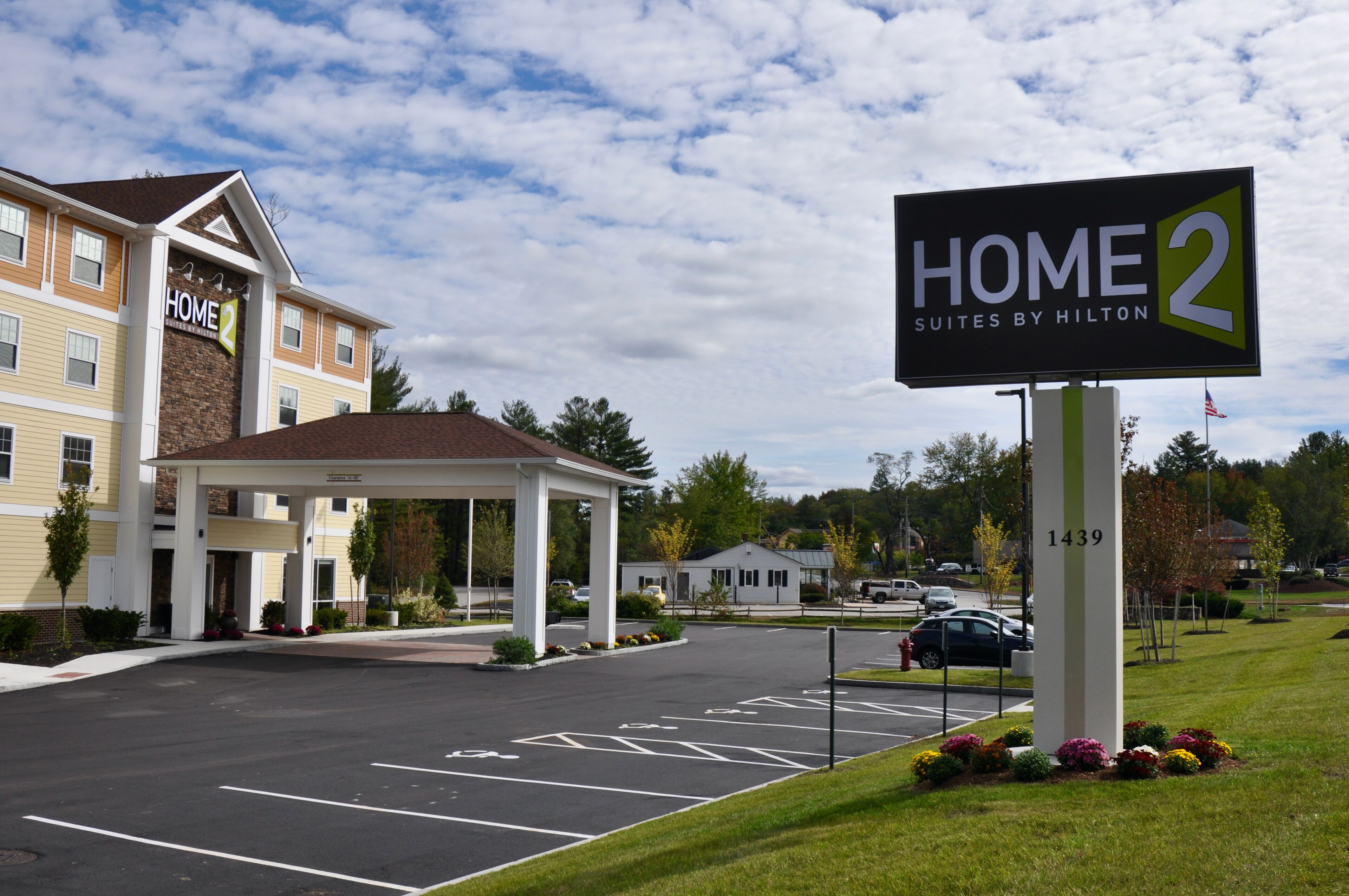 Entrada de hotel com área de espera coberta, placa indicando 'Home 2 Suites by Hilton' e estacionamento.