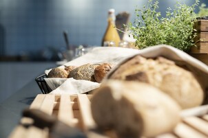 Uma cesta de pão sobre uma mesa, com uma garrafa e uma planta ao fundo.
