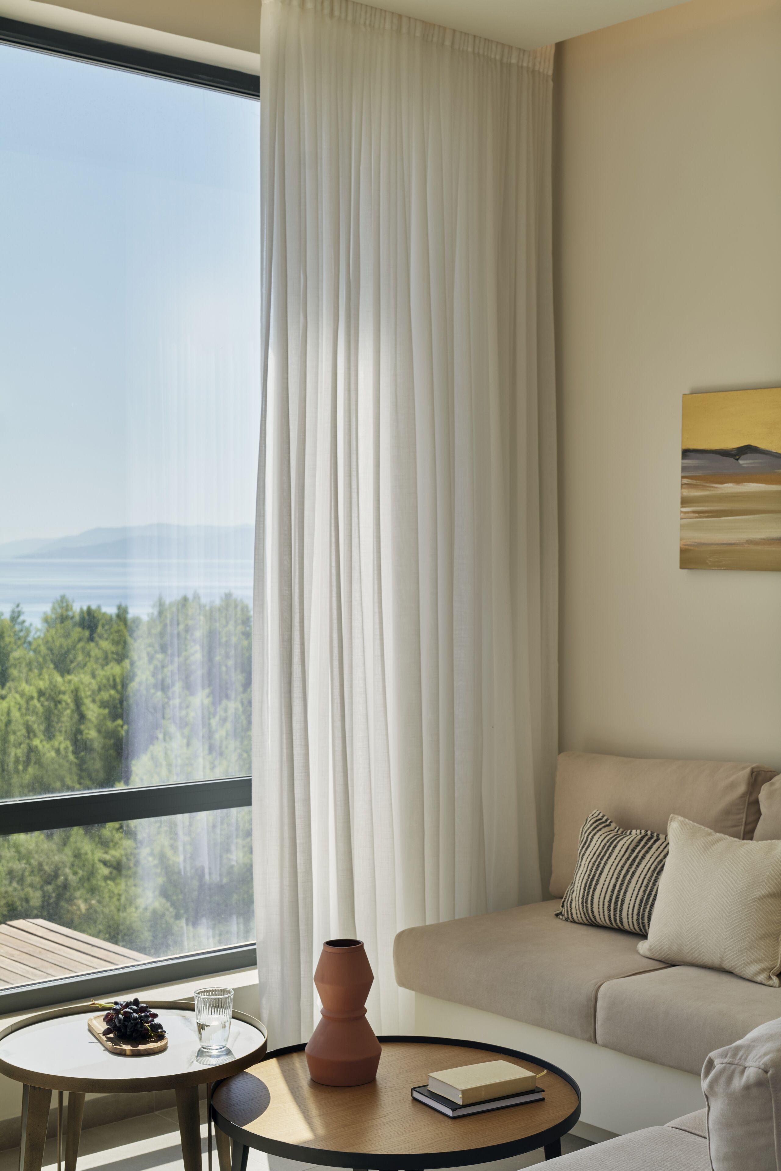 Uma sala de estar moderna com um sofá, uma mesa de centro redonda, uma mesinha com uma tigela de frutas e um copo, e vista para uma floresta através de grandes janelas.
