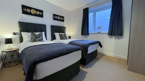 Quarto de hotel com duas camas, uma mesa de cabeceira, uma luminária e uma janela com cortinas.