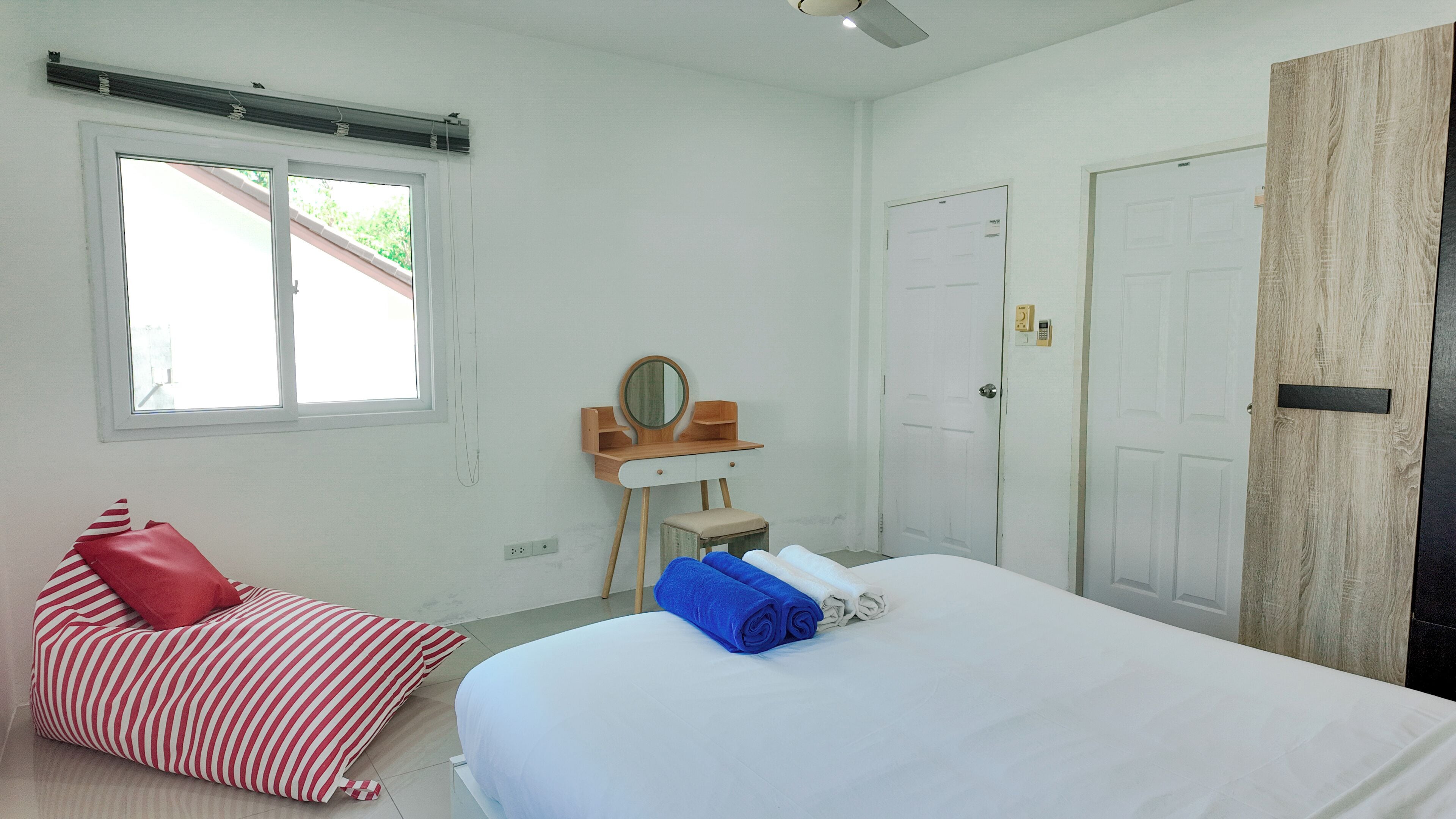 Quarto com cama, mesa de cabeceira com espelho, travesseiro listrado em vermelho e branco, e janela.