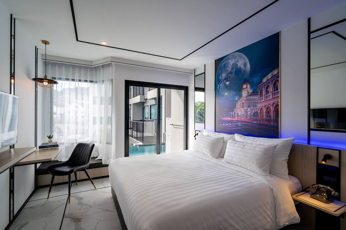 Quarto de hotel moderno com uma cama grande, uma escrivaninha, uma cadeira, uma mesinha de cabeceira e um quadro com uma vista urbana.
