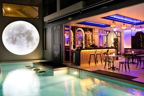 Uma área de piscina interior moderna com uma grande escultura iluminada de lua, um bar com assentos e uma escadaria que dá para um andar superior.