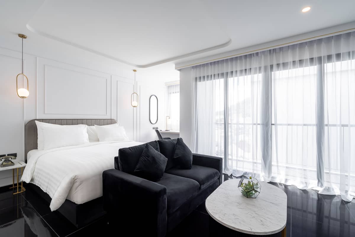 Quarto de hotel moderno com cama, sofá preto, mesa redonda de mármore e cortinas leves.