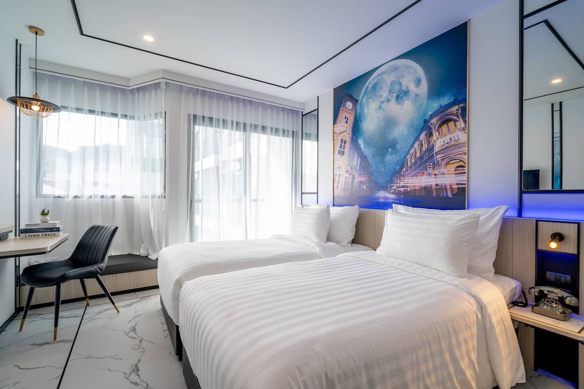 Quarto de hotel com duas camas, uma escrivaninha com cadeira, uma janela grande com cortinas leves e um mural na parede retratando uma paisagem urbana iluminada pela lua.