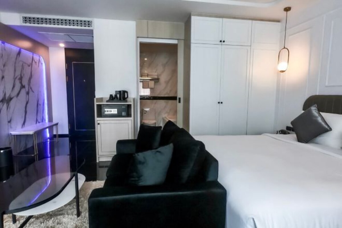 Quarto de hotel moderno com uma cama grande, um sofá preto, uma pequena escrivaninha e uma cozinha compacta ao fundo.