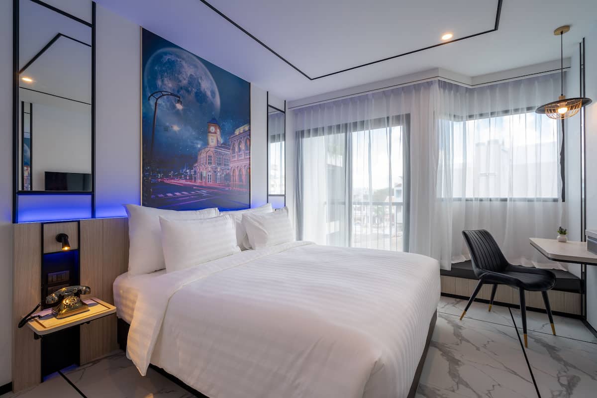 Quarto de hotel moderno com uma cama grande, uma escrivaninha com cadeira, mesa de cabeceira com telefone, uma janela ampla com cortinas leves e um quadro pendurado na parede retratando uma paisagem urbana.