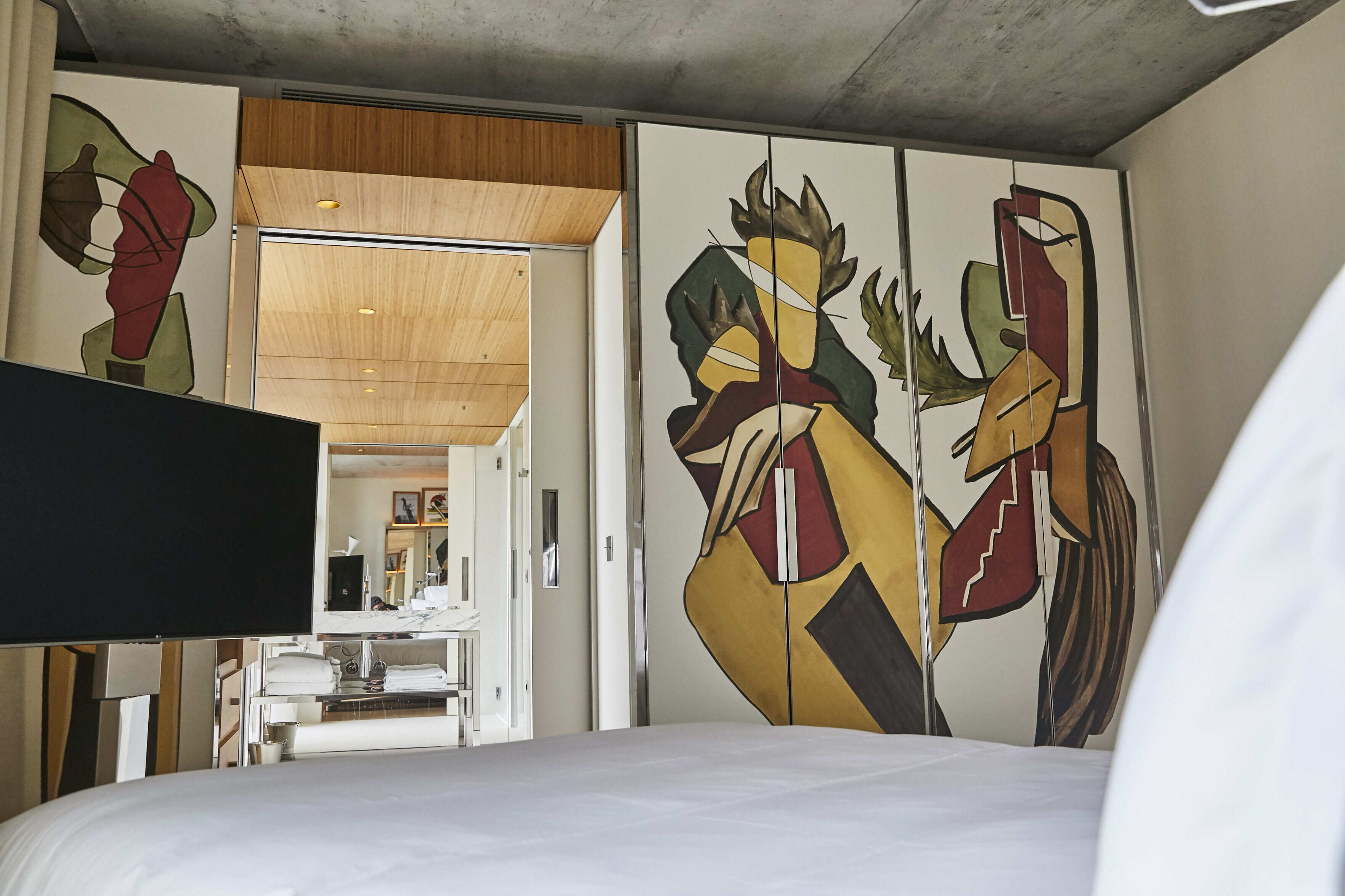Quarto de hotel com cama, televisão, escrivaninha com cadeira e uma grande pintura abstrata na parede.