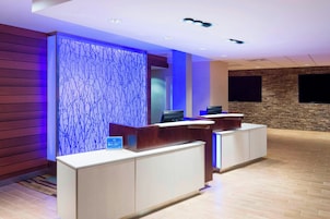 Uma área de recepção com design moderno, destacando-se um balcão de atendimento, uma parede decorativa com iluminação azul, uma parede de pedra e uma televisão instalada.