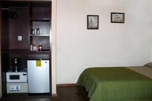 Um quarto pequeno com uma cama, uma cozinha compacta com geladeira e micro-ondas, e duas fotos emolduradas na parede.