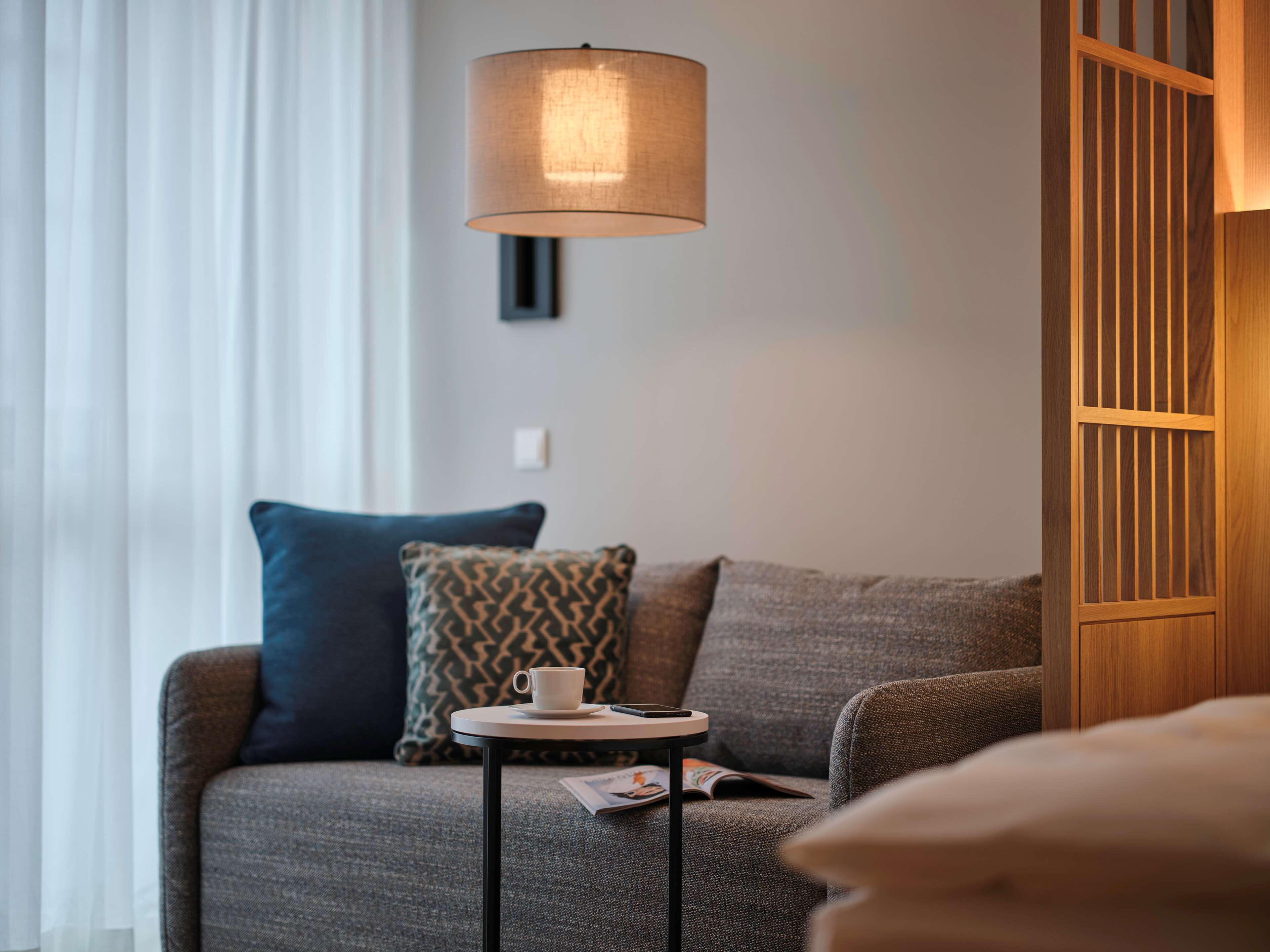 Quarto de hotel moderno com sofá cinza, uma pequena mesa redonda com uma chávena e um comando, e uma janela grande com cortinas brancas.