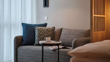 Quarto de hotel moderno com sofá cinza, uma pequena mesa redonda com uma chávena e um comando, e uma janela grande com cortinas brancas.